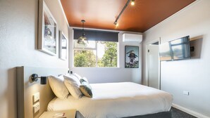Quarto de hotel com cama, mesa de cabeceira, abajur, janela, obras de arte e televisão na parede.