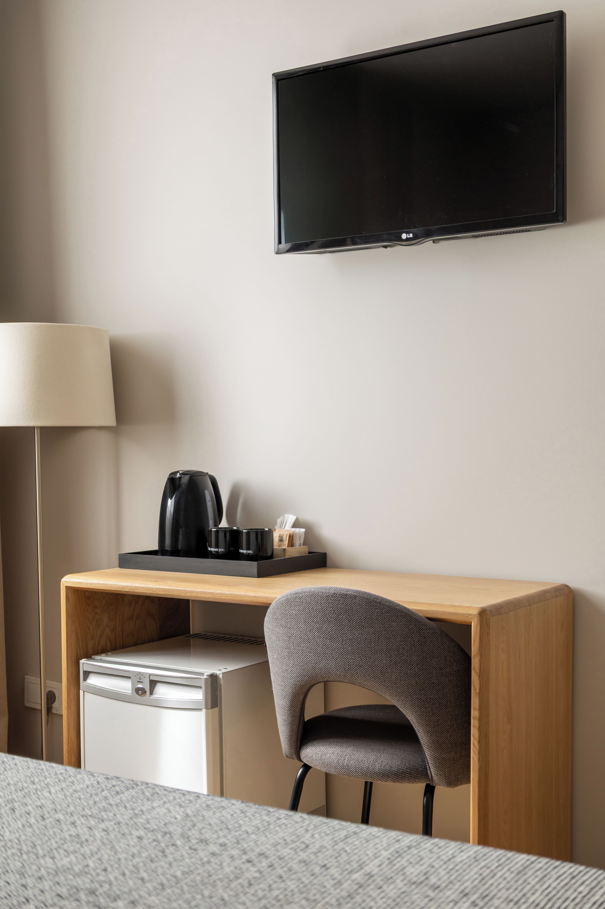 Quarto de hotel compacto com cama, escrivaninha com cadeira, televisão, luminária, cafeteira e pequeno frigorífico.