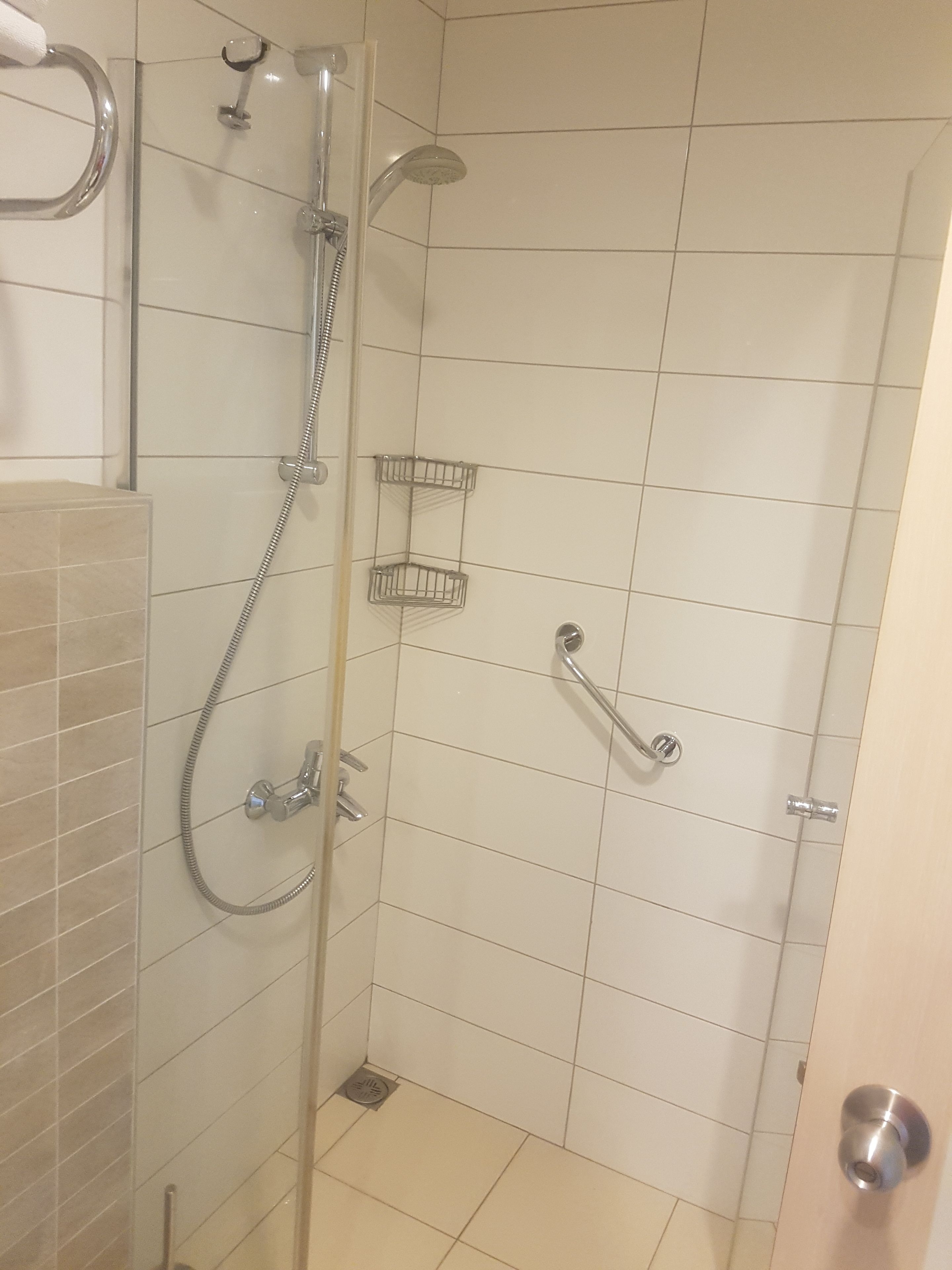 Um chuveiro com porta de vidro, um chuveiro manual, um chuveiro fixado na parede e uma prateleira no canto.