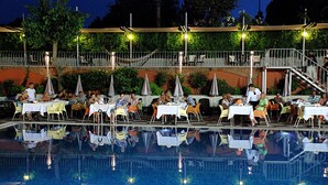 Uma área de refeições ao ar livre, com mesas e cadeiras ao redor de uma piscina, com guarda-sóis e uma escadaria ao fundo.