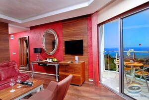 Quarto de hotel com parede vermelha, um espelho grande, televisão de tela plana, escrivaninha de madeira, sofá, mesa de jantar com cadeiras e varanda com vista para o mar.