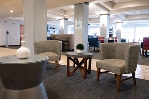 Uma área de recepção com sofás e poltronas, uma pequena mesa com um vaso, e uma placa indicando a localização dos quartos.