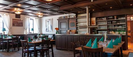 Interior de restaurante com mesas e cadeiras de madeira, área de bar com prateleiras de copos e teto com vigas aparentes.