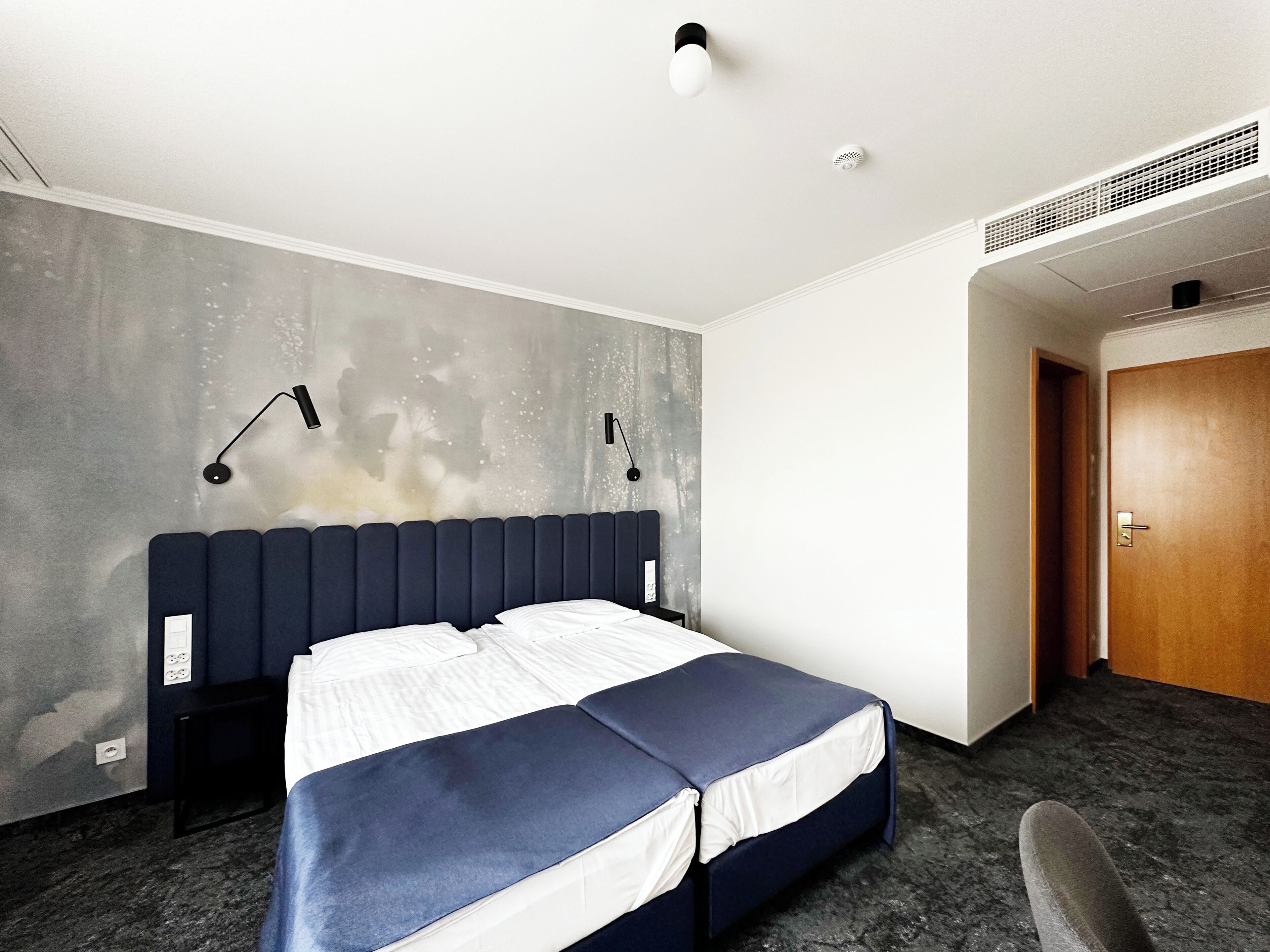 Quarto de hotel com cama, duas luminárias de cabeceira, porta de madeira e parede texturizada.