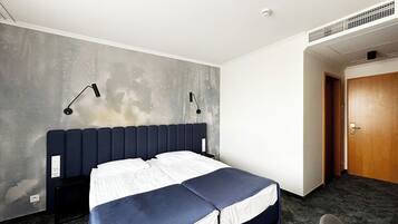 Quarto de hotel com cama, duas luminárias de cabeceira, porta de madeira e parede texturizada.