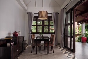 Uma sala de jantar com mesa e cadeiras, luminária pendente, janela com cortinas e vista para o verde lá fora.