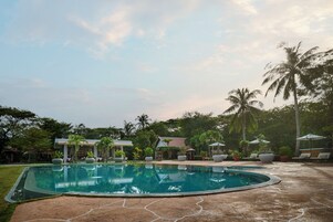 Piscina com espreguiçadeiras e guarda-sóis, rodeada por palmeiras e um edifício com telhado vermelho.