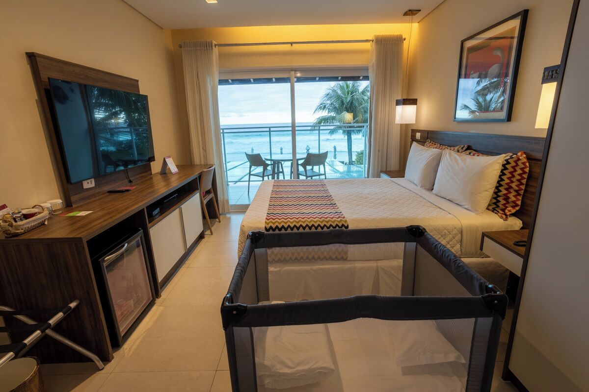 Quarto de hotel com cama, escrivaninha com televisão, varanda e vista para o mar, e uma cadeira.