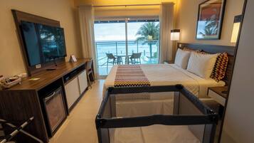 Quarto de hotel com cama, escrivaninha com televisão, varanda e vista para o mar, e uma cadeira.