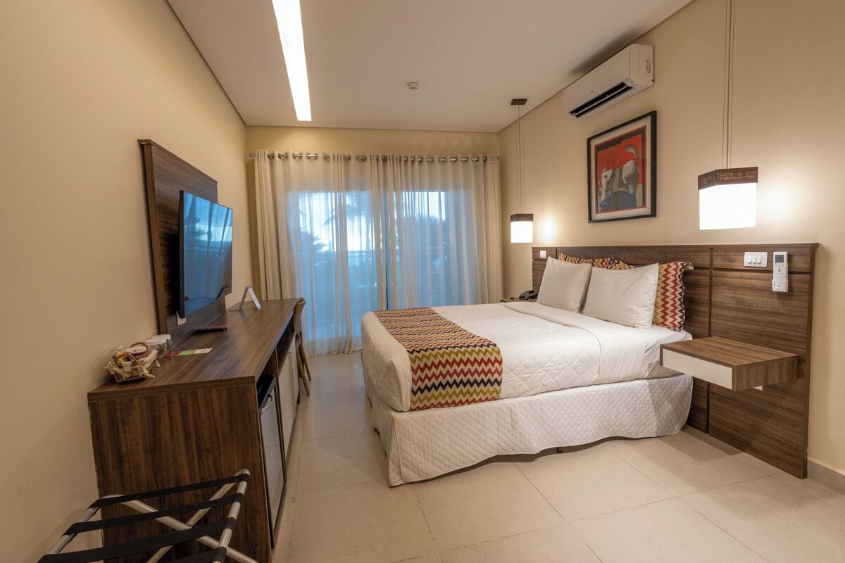 Quarto de hotel com cama, mesa de cabeceira, televisão e cortinas.