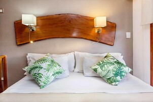 Cama com roupa de cama branca e duas almofadas com padrão de folhas verdes, cabeceira de madeira com luzes fixadas na parede e uma parede lisa.