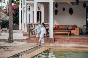 Um casal ao lado de uma piscina com deck de madeira, um edifício branco com um banco e paisagem tropical.
