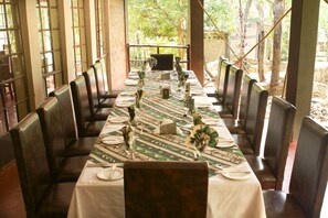 Uma longa mesa de jantar posta com cadeiras, toalha de mesa estampada e peças decorativas no centro.
