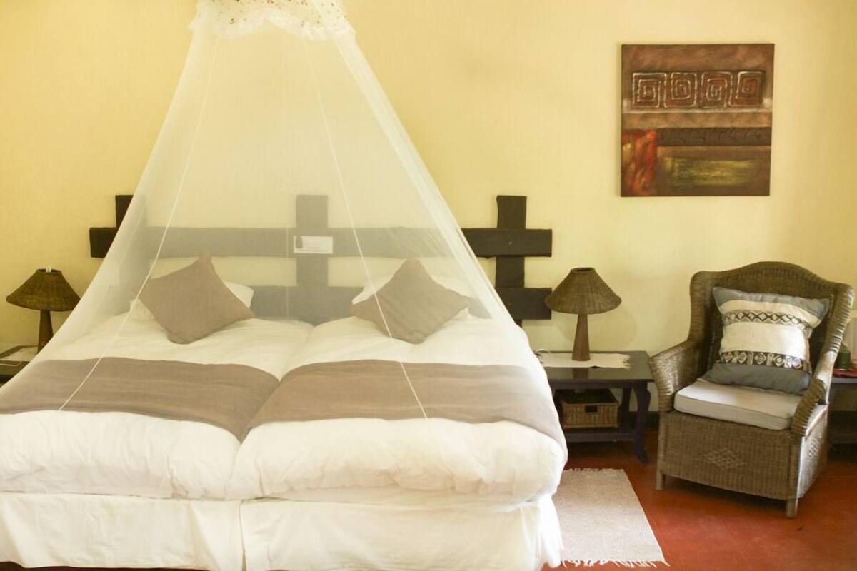 Quarto com cama de dossel, duas mesas de cabeceira com abajures, uma cadeira e uma cruz na parede.