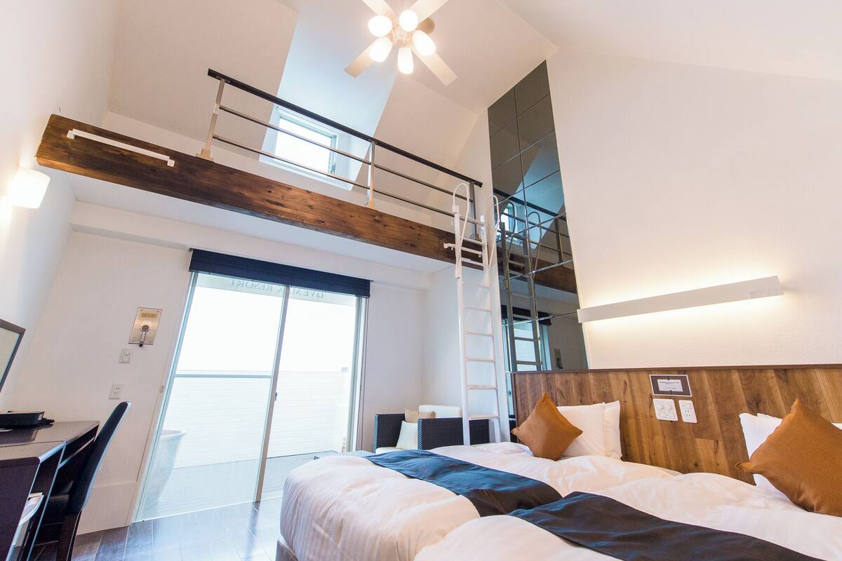 Quarto de hotel moderno com área de dormir elevada, uma escrivaninha e uma janela grande.