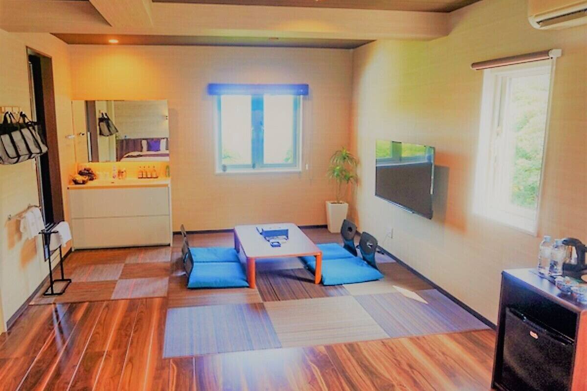 Quarto compacto com cama, uma mesinha, televisão e cozinha compacta.