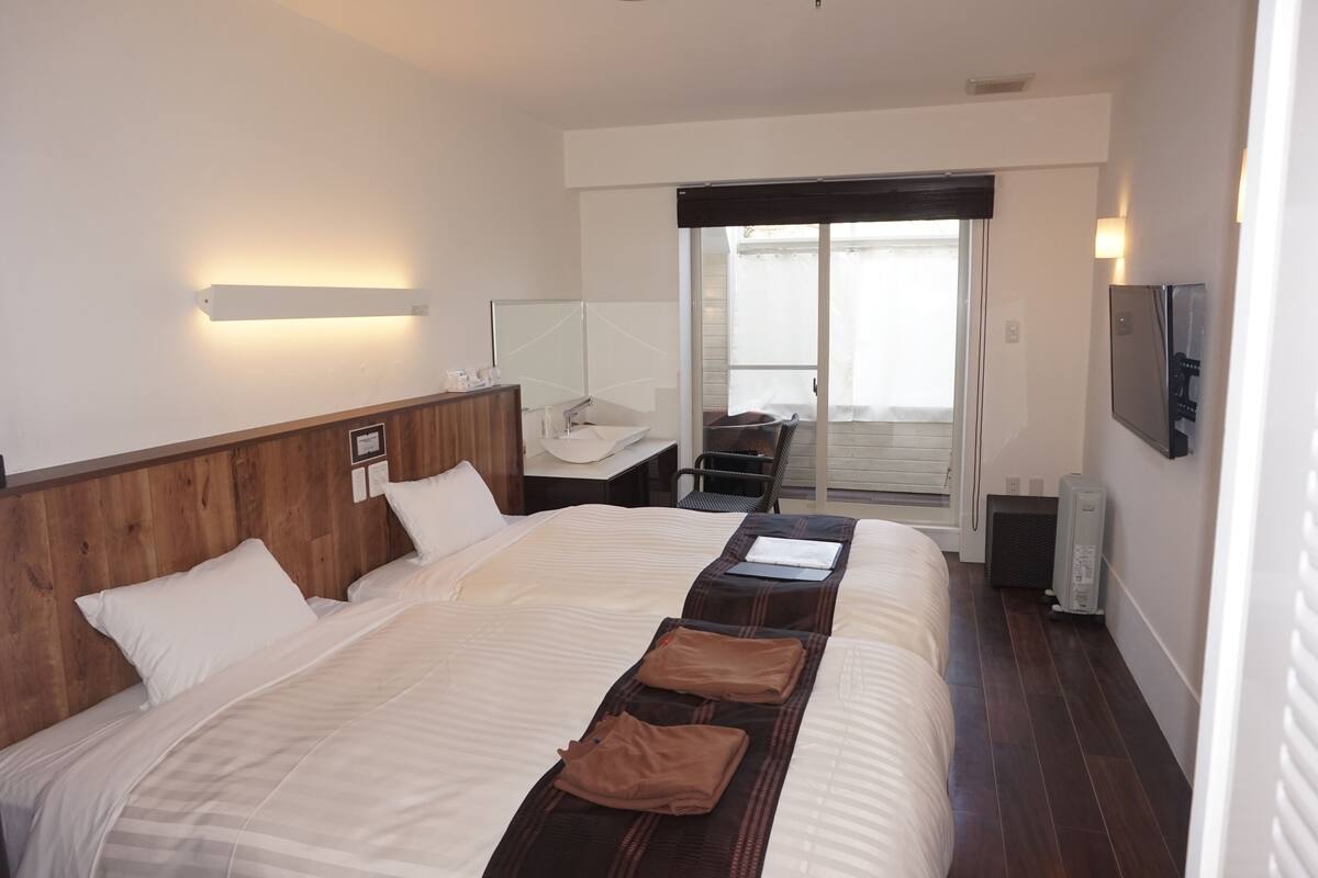 Quarto de hotel com uma cama grande, uma escrivaninha com cadeira, uma porta de vidro deslizante para uma varanda e uma TV fixada na parede.