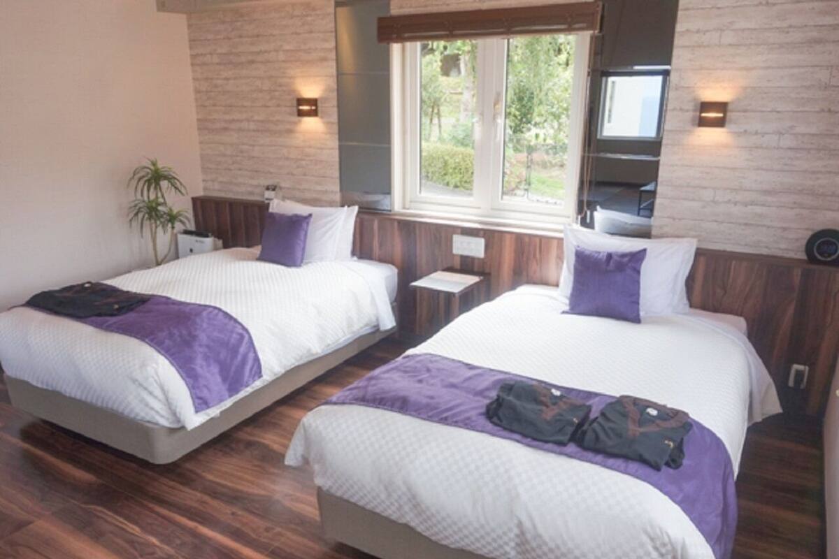 Quarto de hotel com duas camas, uma mesinha de cabeceira, televisão e janela com vista para áreas verdes.