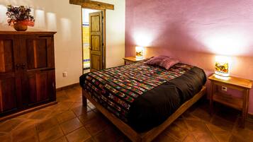 Quarto com cama de madeira, mesa de cabeceira com abajur, guarda-roupa de madeira e porta para outro cômodo.
