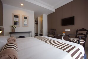 Quarto de hotel com uma cama grande, duas cadeiras, uma mesinha com um vaso de flores, uma TV de tela plana na parede e uma porta que dá para outro quarto.