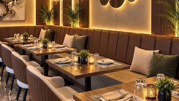 Interior moderno de restaurante com uma longa mesa de jantar, assentos acolchoados e iluminação ambiente.