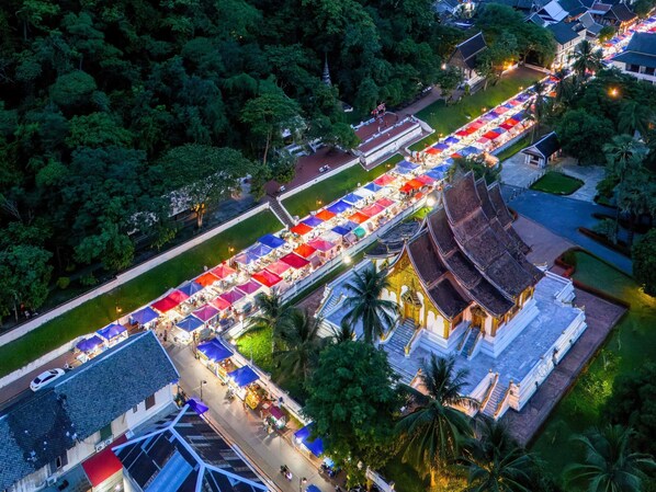 Vista aérea de uma rua repleta de bancas de mercado coloridas, um templo tradicional e vegetação ao redor.