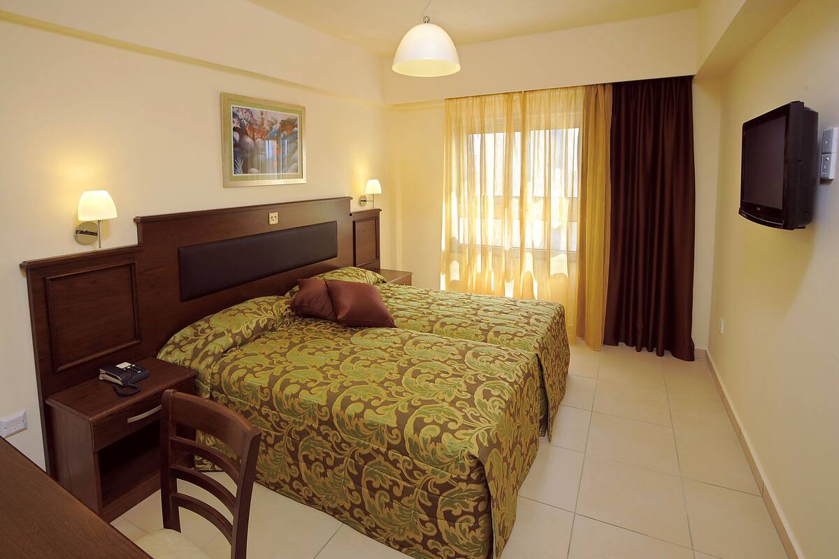 Quarto de hotel com cama, mesas de cabeceira, escrivaninha, cadeira, televisão e janela com cortinas.