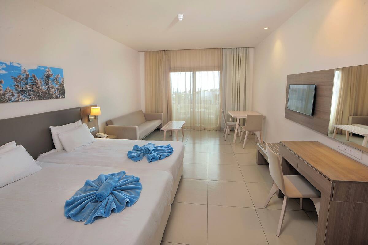 Quarto de hotel com duas camas, uma escrivaninha, uma cadeira, uma televisão e um quadro na parede.