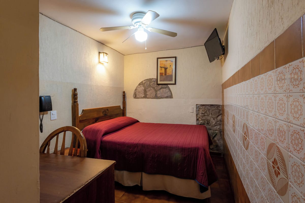 Quarto pequeno e acolhedor com uma cama de solteiro, uma cadeira de madeira, uma escrivaninha, um ventilador de teto, televisão na parede e uma parede de detalhes em pedra.