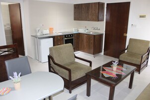Uma cozinha compacta com eletrodomésticos modernos, duas cadeiras de madeira, uma mesa de centro de vidro e uma pequena mesa de jantar com um vaso de planta.