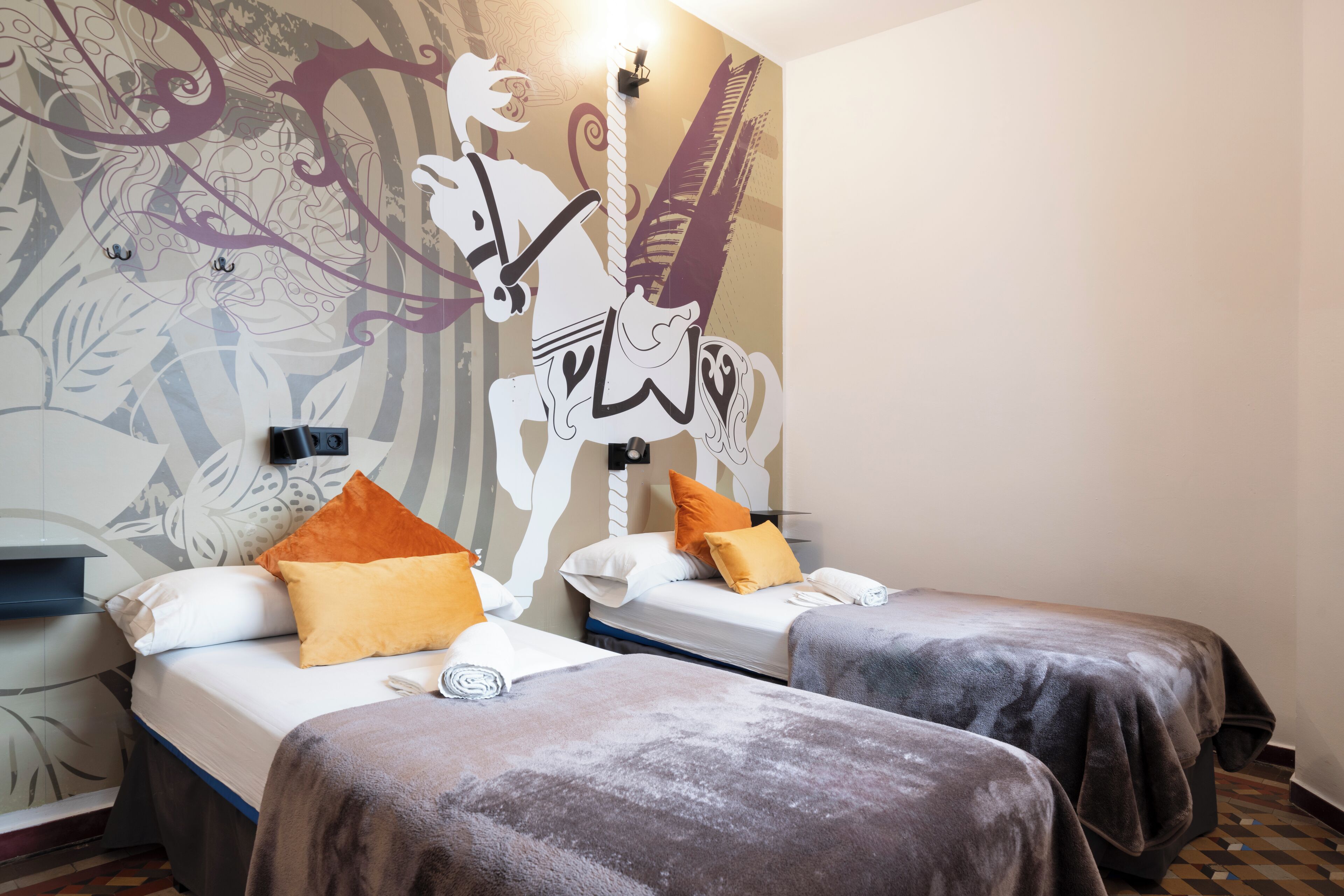 Quarto com duas camas, cada uma com manta cinza e almofadas laranja. Há um mural na parede com um cavalo e um cavaleiro.