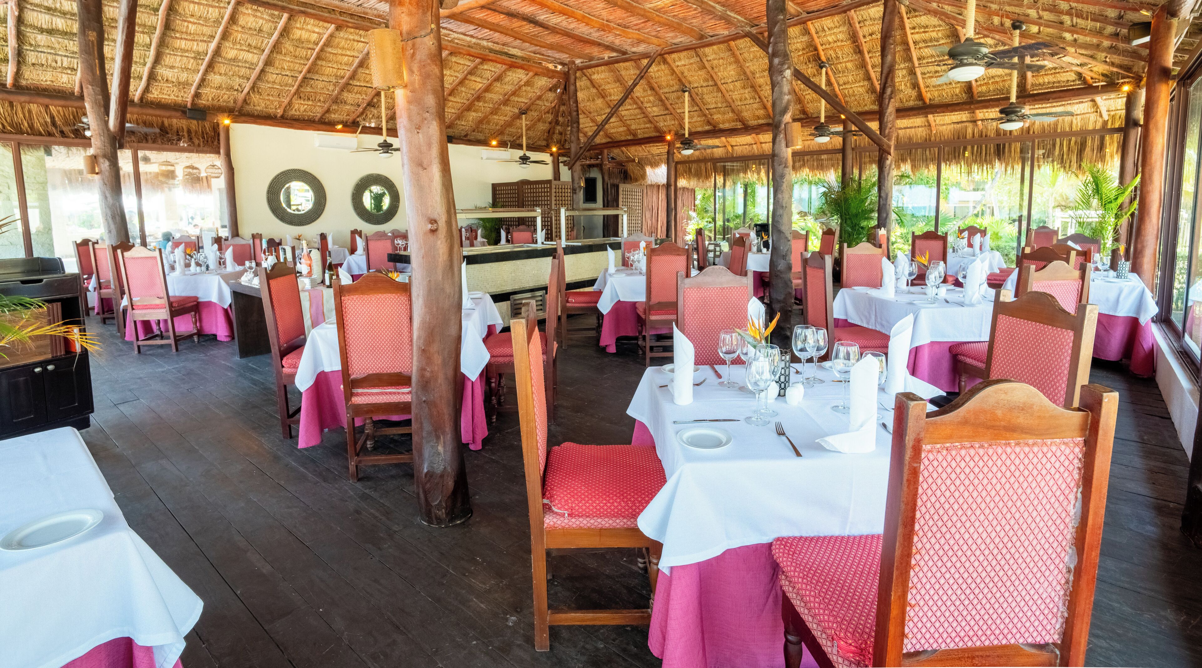 Um restaurante ao ar livre com mesas e cadeiras de madeira, telhado de palha e luz natural.