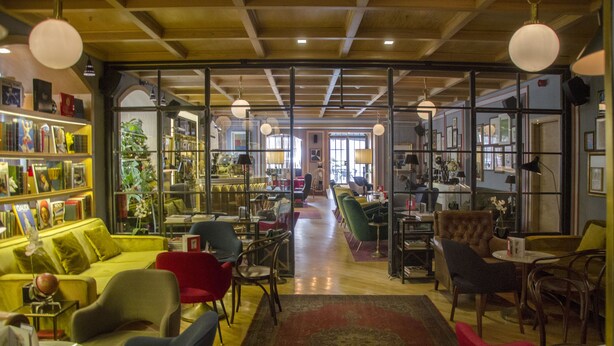 Interior espaçoso com uma passagem central que leva a uma área de bar, rodeada por diversas opções de assento e uma estante cheia de livros.