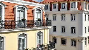Um edifício de vários andares com telhado de telhas vermelhas, fachadas brancas e amarelas, e varandas de ferro forjado preto.