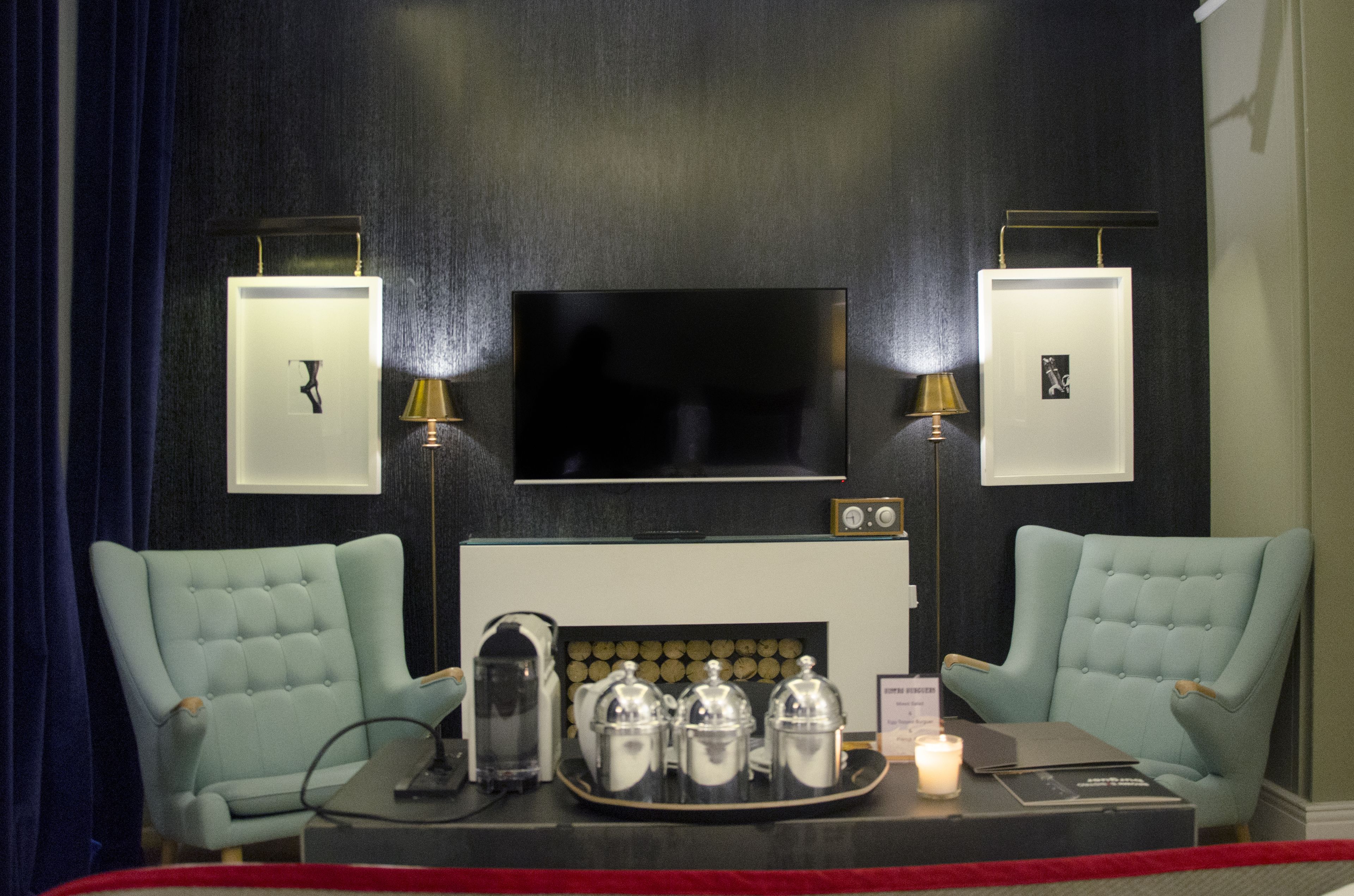 Quarto com duas poltronas azul claras, uma mesinha com uma vela, um tabuleiro com recipientes de vidro e uma TV de tela plana fixada na parede.