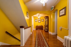 Um corredor com paredes amarelas, piso de madeira, tapete vermelho estampado, ventilador de teto e quadros nas paredes.