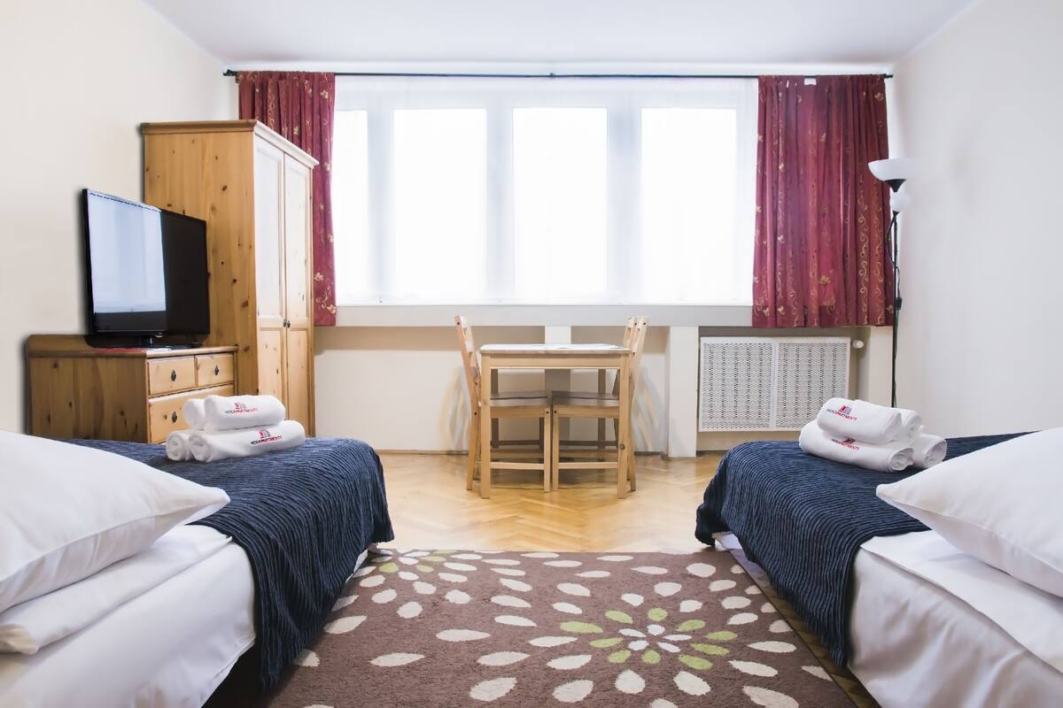 Quarto de hotel com duas camas, televisão, aparador de madeira, mesa e janela com cortinas.