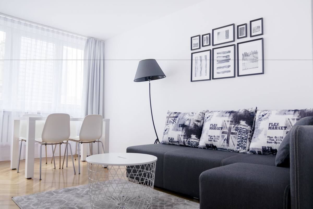 Uma sala de estar moderna com um sofá cinza, cadeiras brancas, uma mesa de centro redonda e um abajur de chão preto. Há quadros nas paredes e uma janela com cortinas finas.
