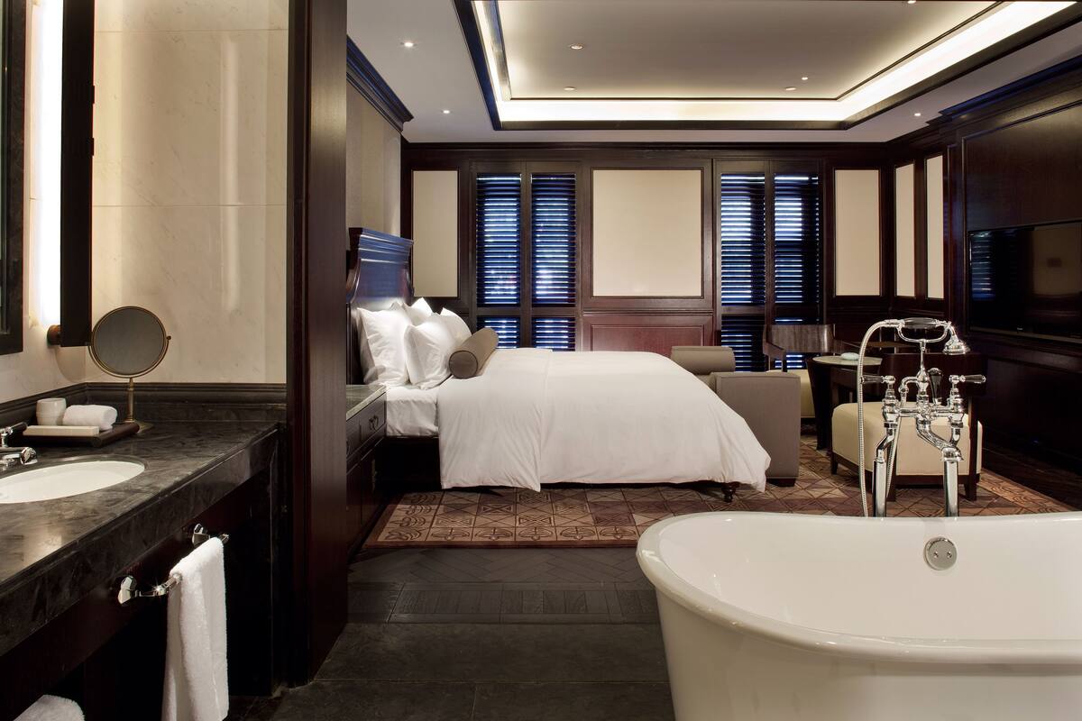 Quarto de hotel moderno com uma cama grande, área de estar separada, banheiro com banheira e um espelho amplo.