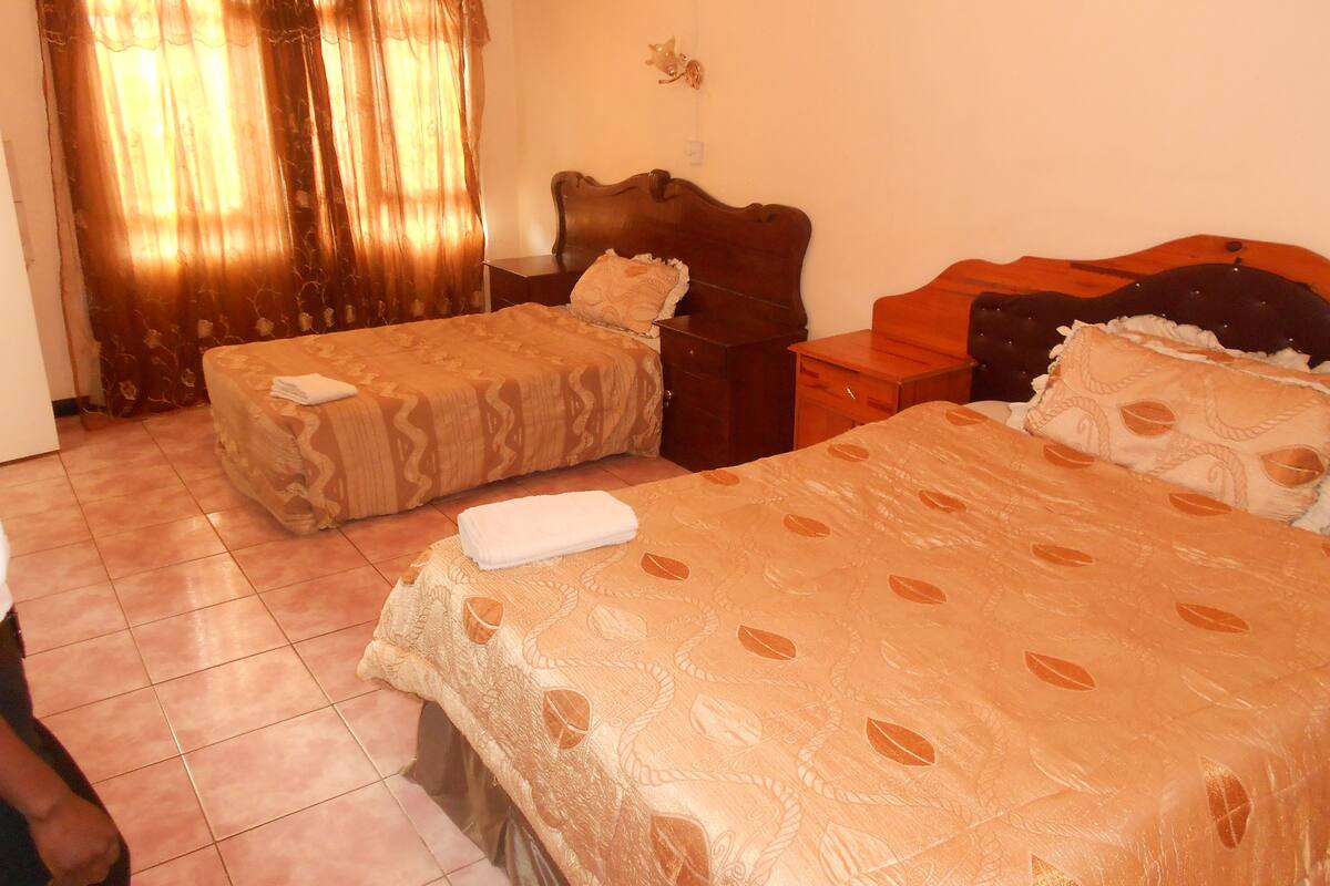 Quarto de hotel com duas camas, cabeceira de madeira, mesa de cabeceira e cortina.