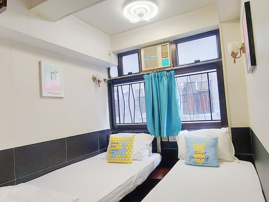 Quarto de hotel com duas camas de solteiro, janela com cortinas, ar condicionado fixado na parede e luminária no teto.