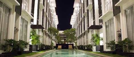 Uma área de piscina com um elemento aquático central, ladeada por edifícios simétricos com janelas amplas e plantas em vasos.