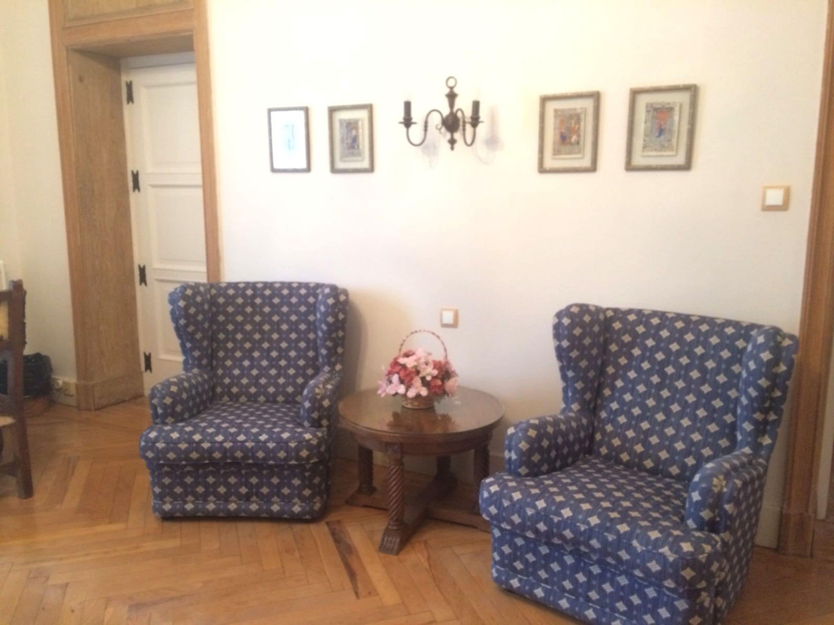 Quarto com duas poltronas estampadas, uma pequena mesa redonda com um vaso de flores e um candelabro fixado na parede.