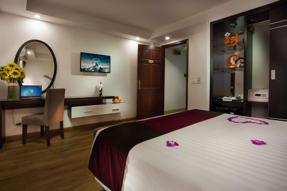 Quarto de hotel com cama, escrivaninha com computador, cadeira, guarda-roupa e televisão na parede.