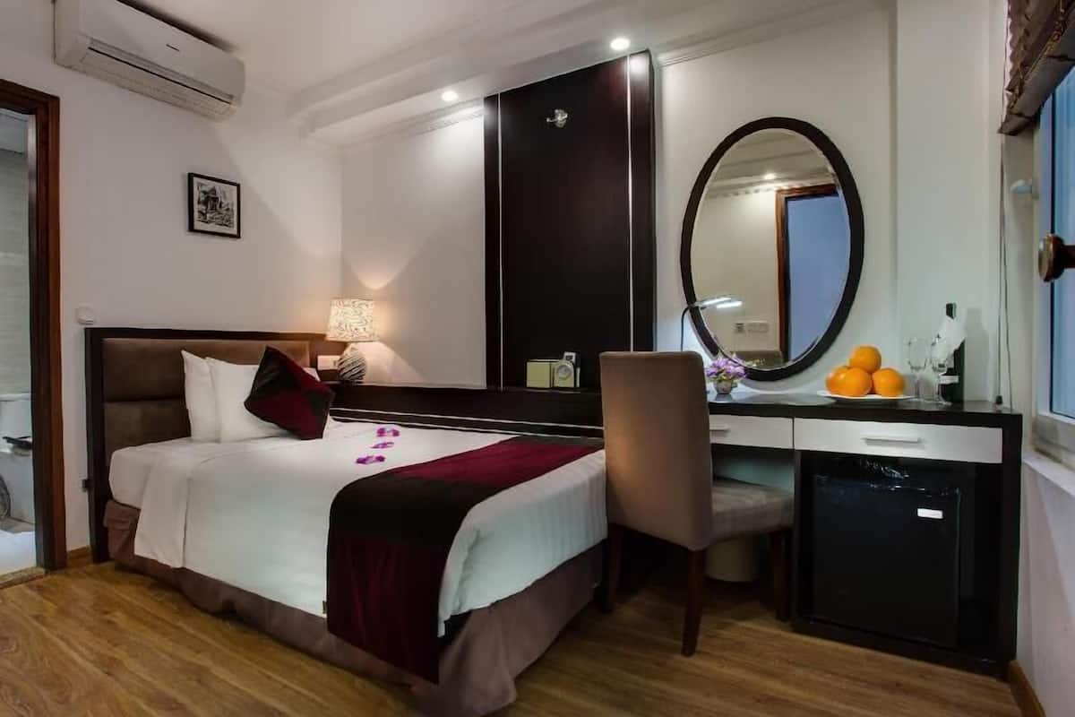 Quarto de hotel com cama, escrivaninha, cadeira, espelho e um pequeno frigorífico.