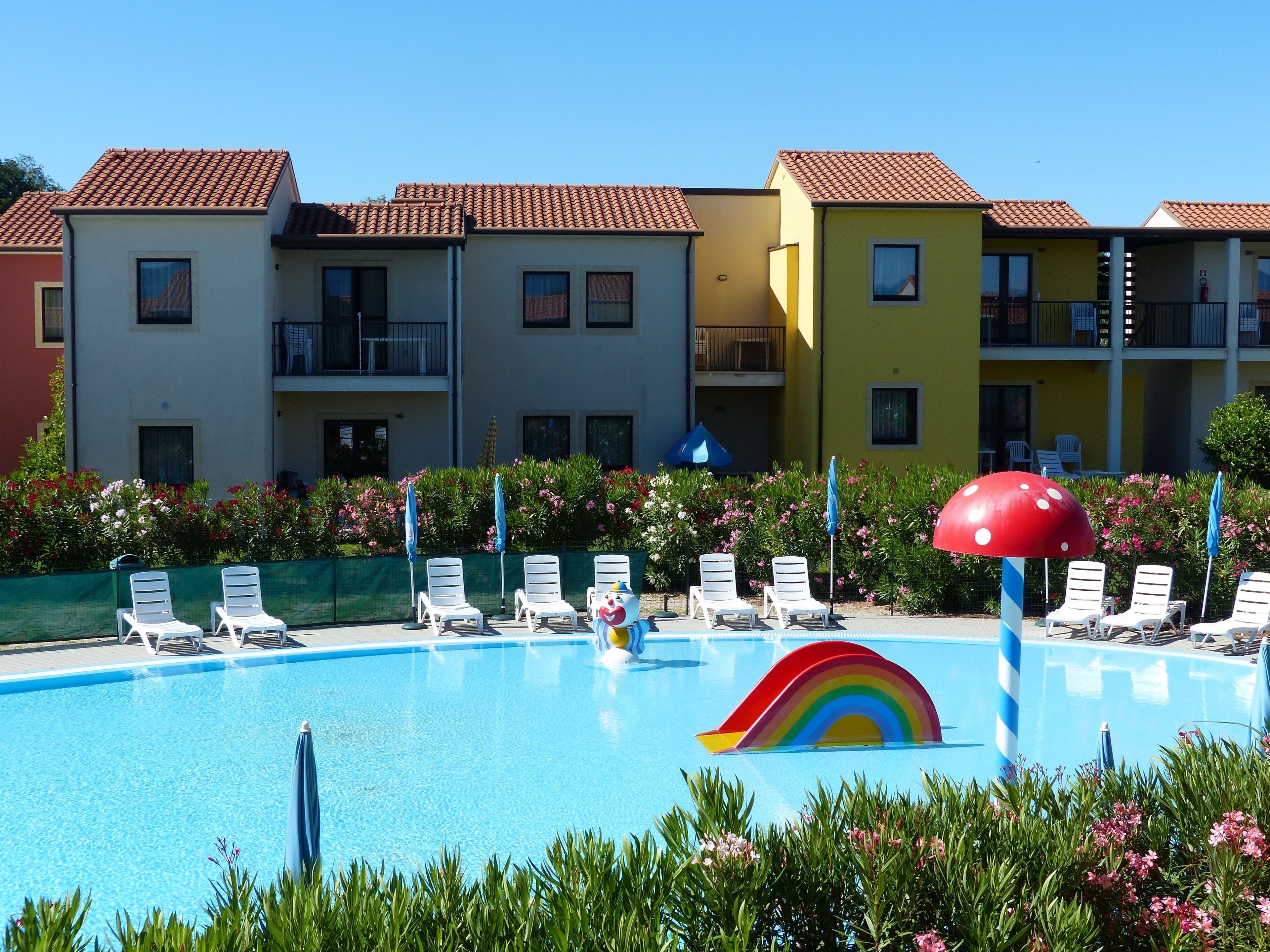 Piscina com espreguiçadeiras e guarda-sóis, rodeada por edifícios coloridos com varandas.