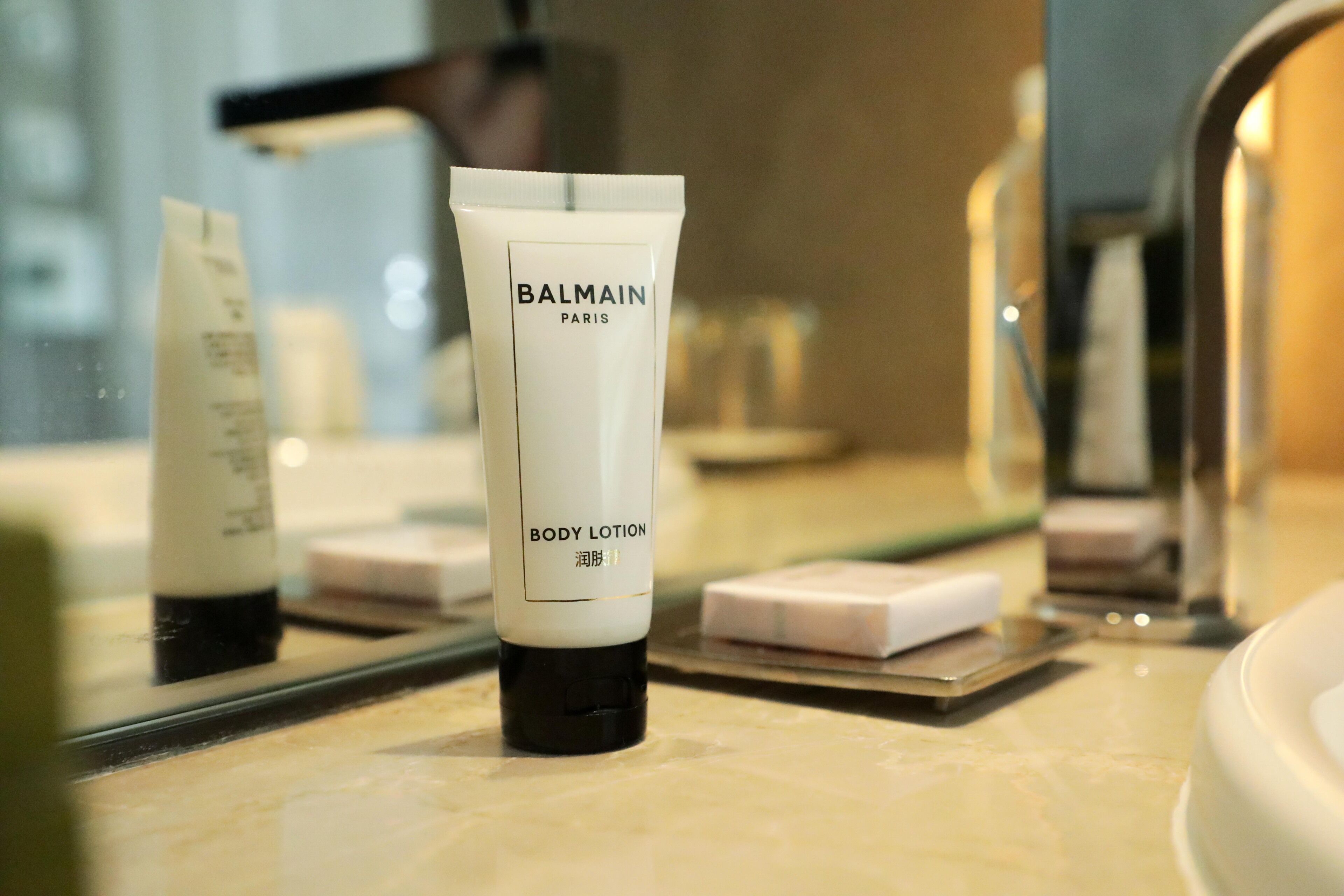 Embalagem de loção corporal Balmain sobre uma bancada de banheiro, com um espelho refletindo uma pia e produtos de higiene pessoal.