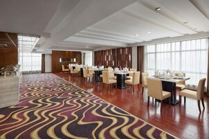 Uma ampla sala de jantar de hotel com v árias mesas e cadeiras, um tapete estampado e grandes janelas.