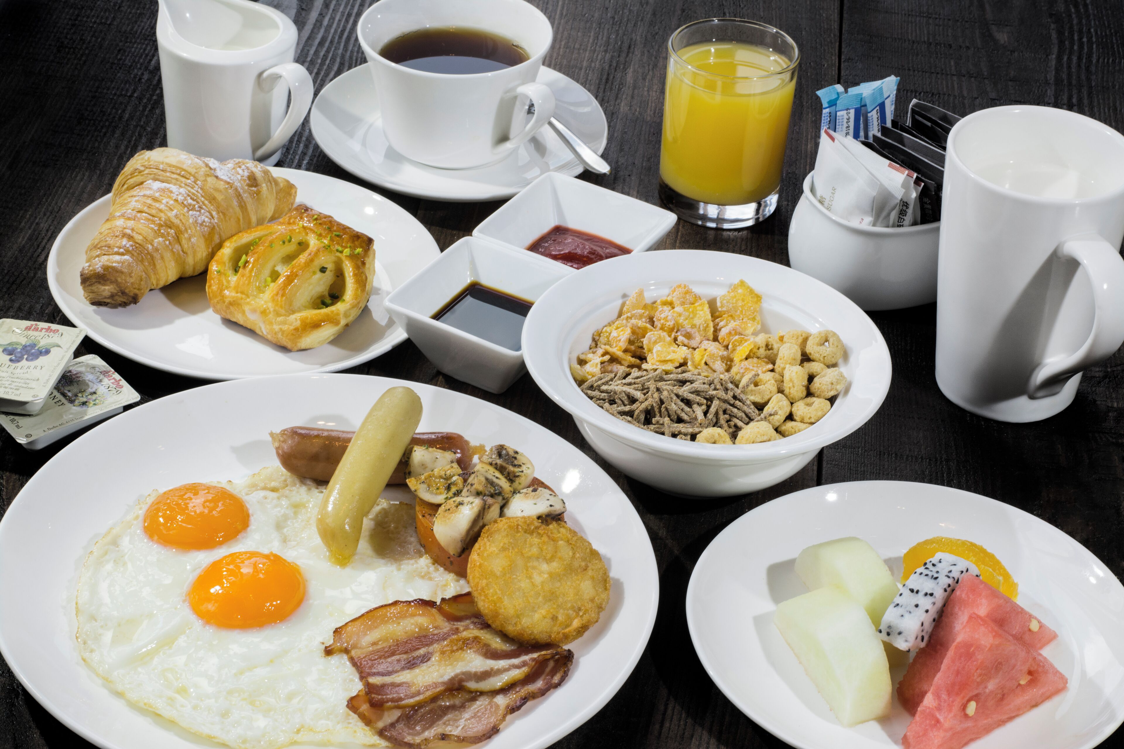 Uma mesa farta com croissants, ovos, bacon, salsichas, frutas variadas e cereais.