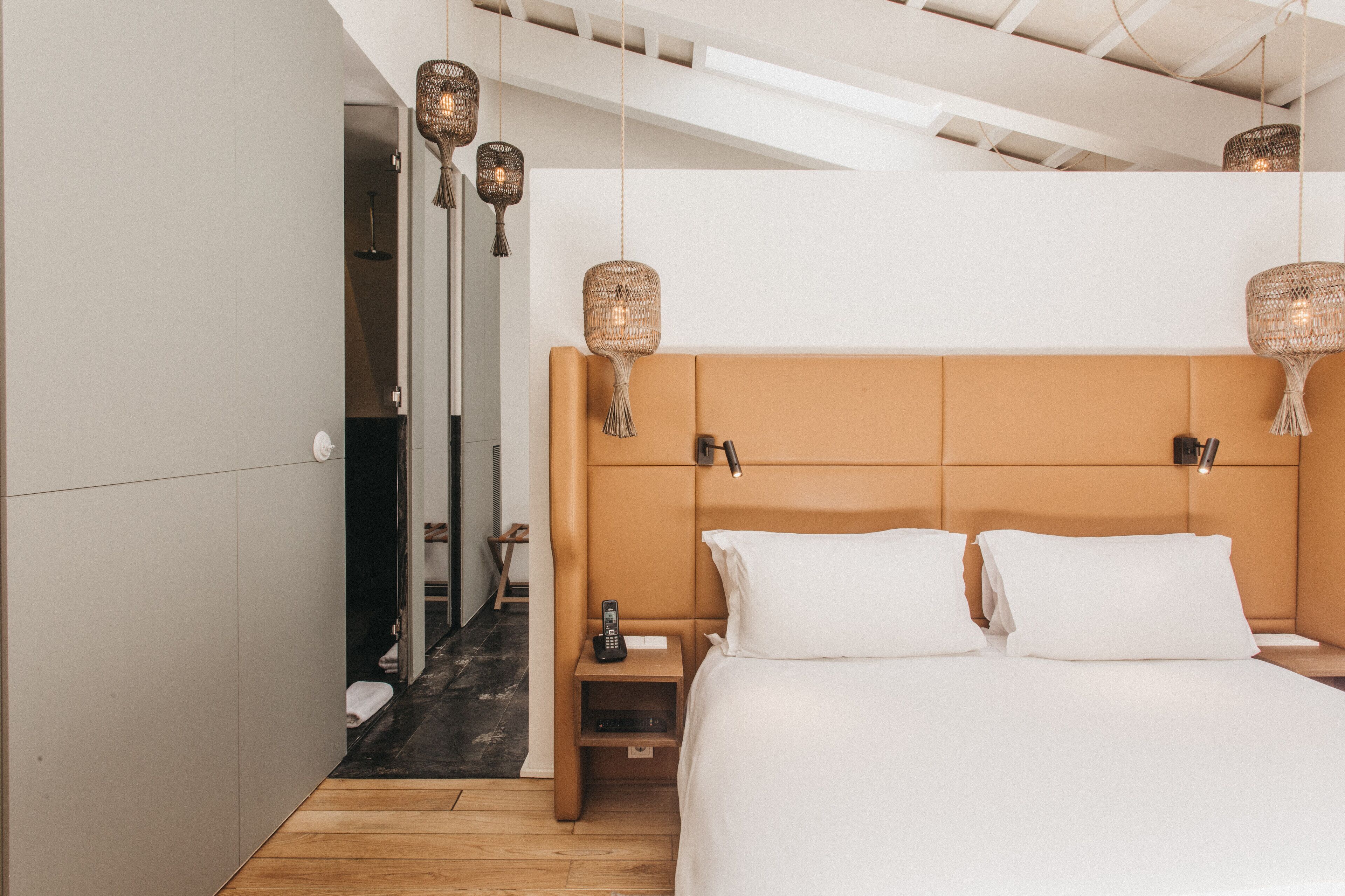 Quarto de hotel moderno com uma cama grande, piso de madeira e uma pequena mesa de cabeceira com telefone.
